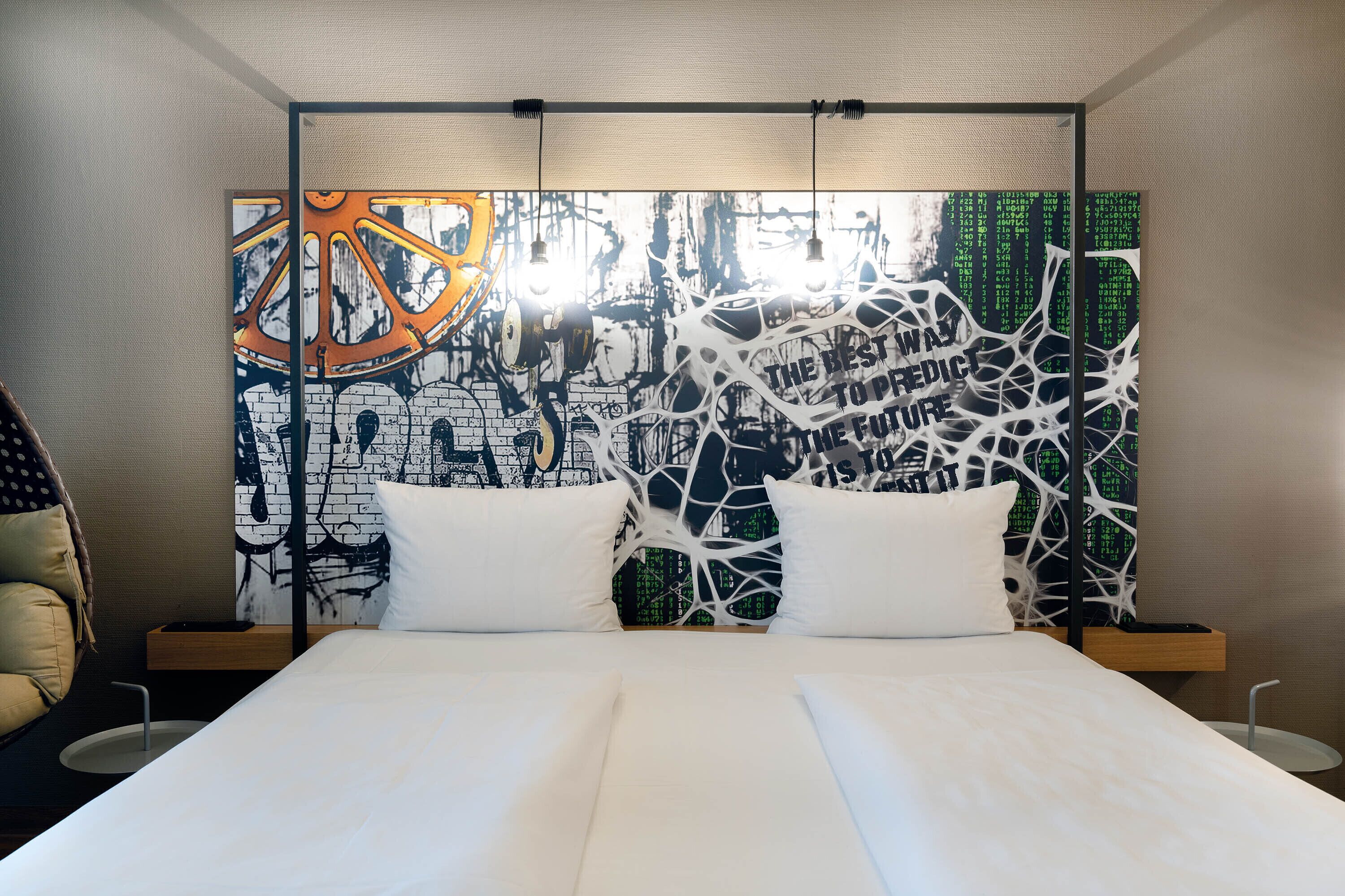 Quarto de hotel moderno com uma cama grande, duas almofadas brancas, cabeceira com arte abstrata e mesa de cabeceira com abajur.