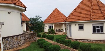 Uma área residencial com três casas, cada uma com telhado de telhas vermelhas e paredes brancas. Há um jardim bem cuidado, com arbustos aparados e um muro de pedra.