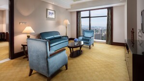 Quarto de hotel com sofá, poltrona e uma mesinha com abajur. Há uma janela grande com vista para a cidade.