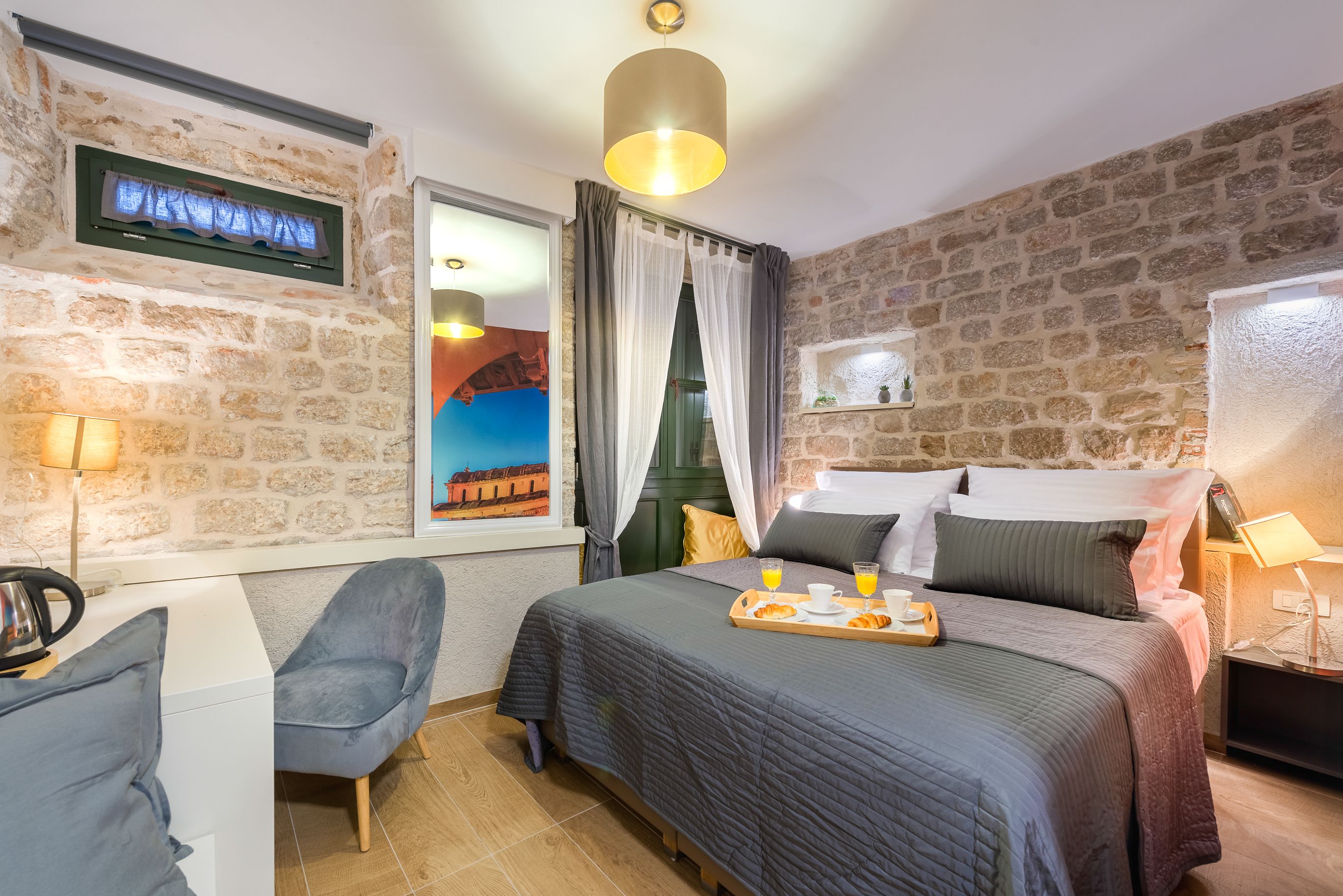 Quarto de hotel acolhedor com parede de pedra, cama com bandeja de café da manhã, uma cadeira e escrivaninha com luminária.