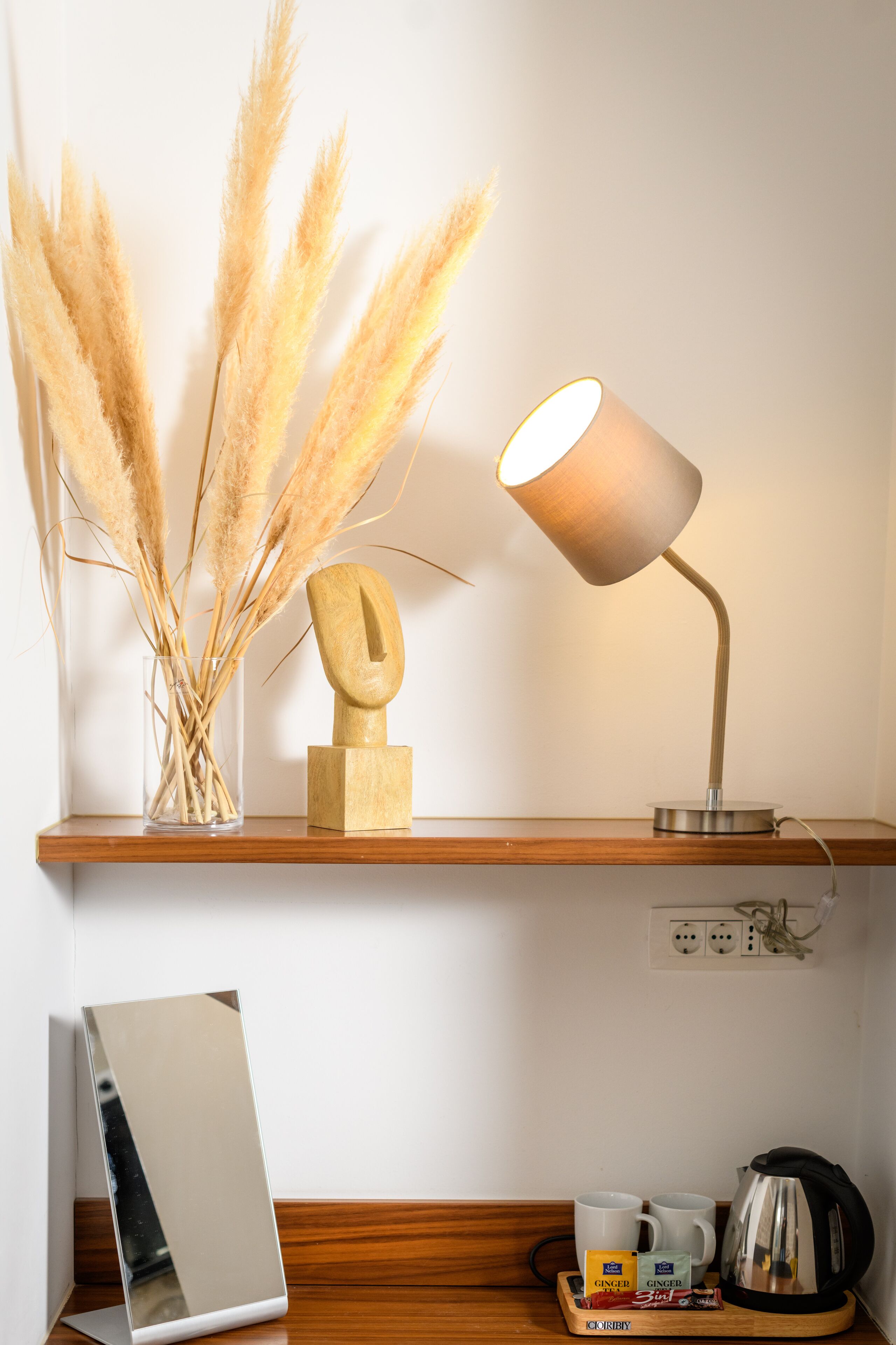 Uma mesa com um laptop, um espelho, uma luminária, um vaso com plumas de capim-do-campo, uma escultura de madeira e uma bandeja com chaleira e xícaras.