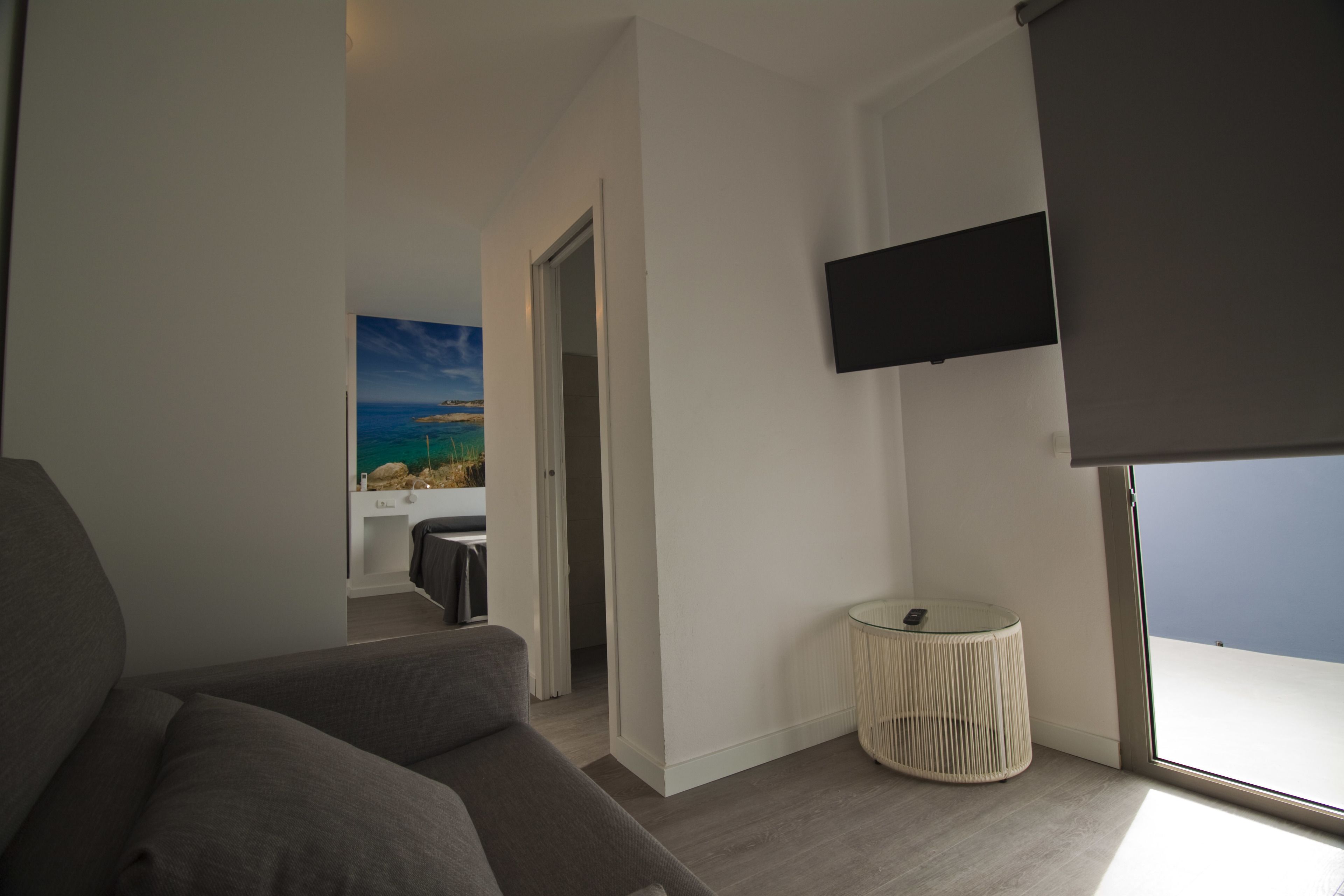 Uma sala de estar moderna com um sofá cinza, uma televisão de tela plana na parede e uma janela grande com vista para o exterior.