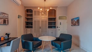 Um quarto acolhedor com uma mesa redonda, duas poltronas azuis, uma máquina de café e uma estante de livros.
