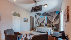 Quarto de hotel moderno com uma cama, dois poltronas, uma mesinha redonda, uma escrivaninha com um computador e uma TV na parede.