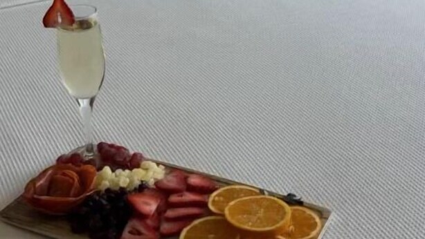 Cama bem arrumada com lençóis brancos, mesa de cabeceira de madeira com abajur, e bandeja com frutas e bebida sobre a cama.