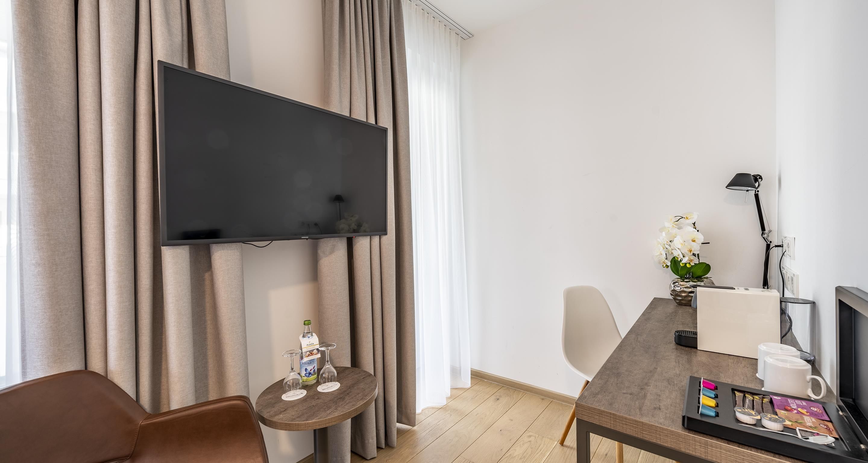 Uma sala de estar moderna com uma TV de tela plana fixada na parede, uma mesa de madeira redonda com um porta-trejos de canetas e um copo, e uma cadeira.
