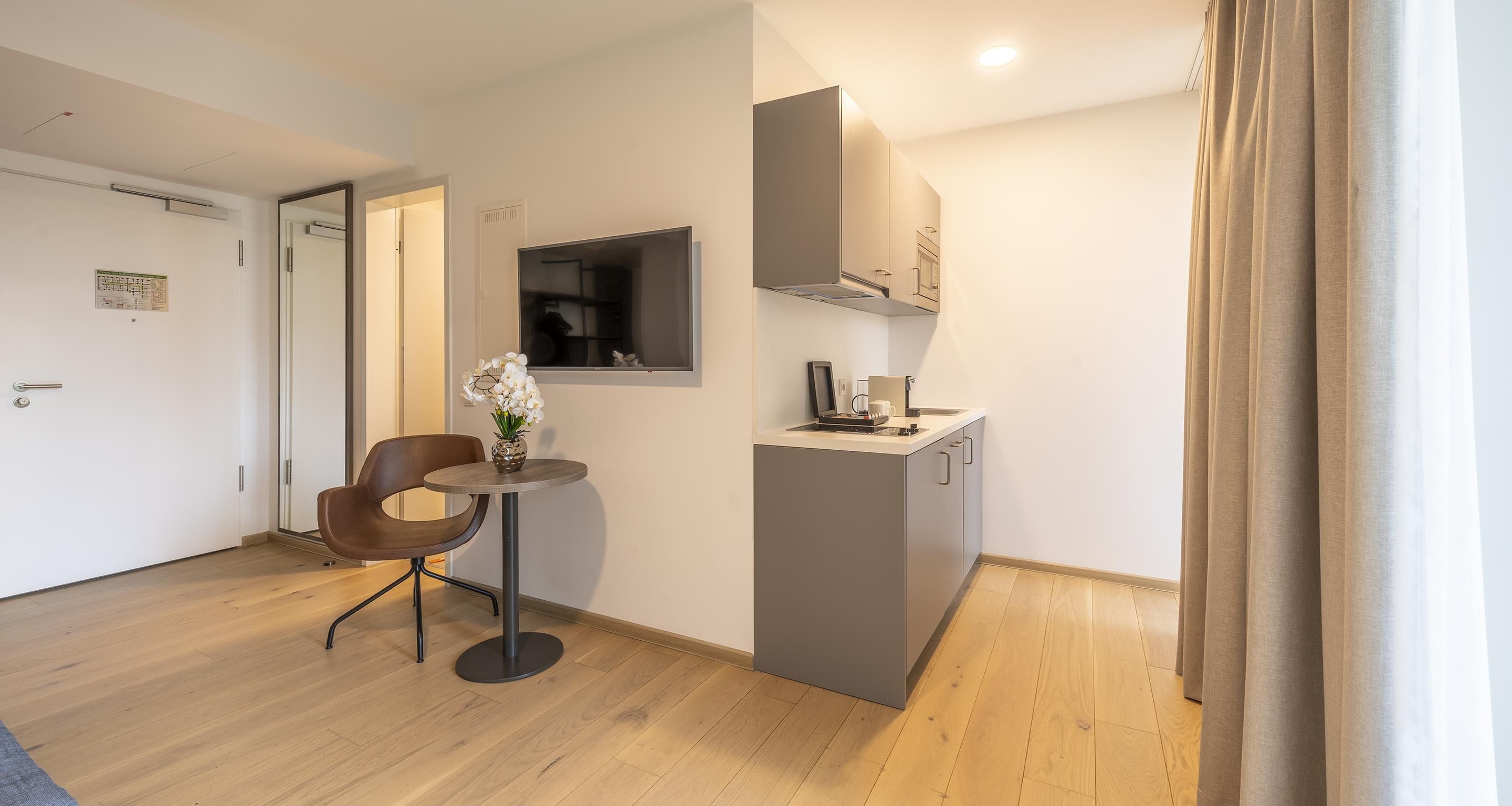 Uma cozinha moderna com forno embutido, uma pequena mesa de jantar com uma cadeira e uma janela com cortinas.