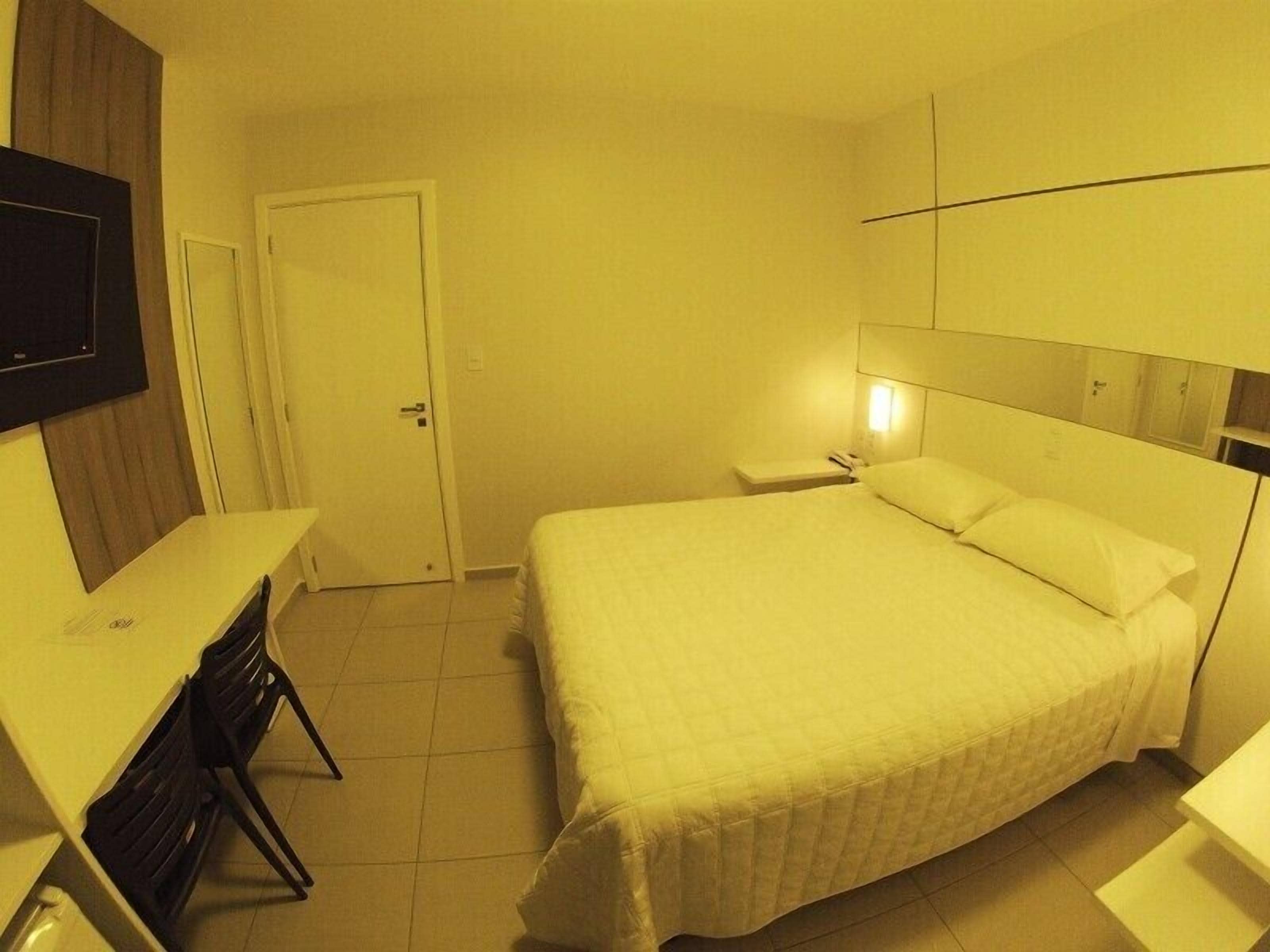 Quarto de hotel com cama, escrivaninha e cadeira. Há uma porta e uma mesa de cabeceira com abajur.