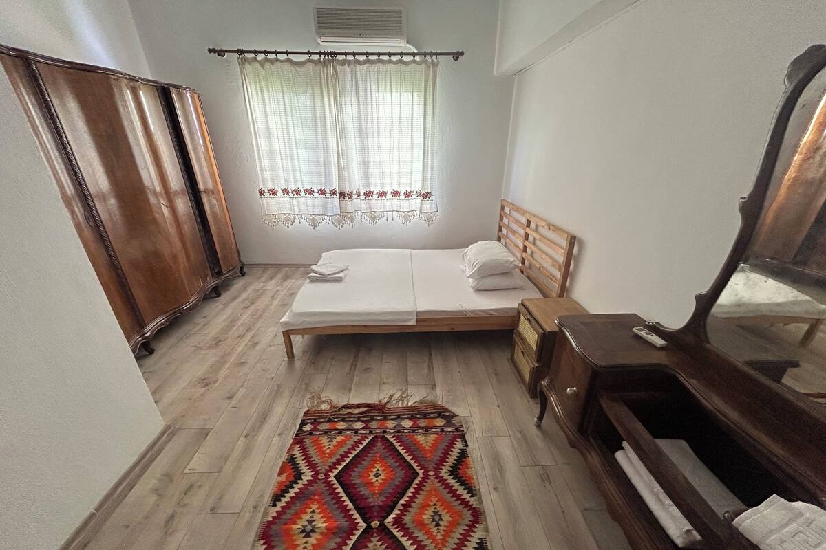 Quarto com cama de madeira, mesa de cabeceira pequena, espelho, guarda-roupa de madeira e tapete estampado.