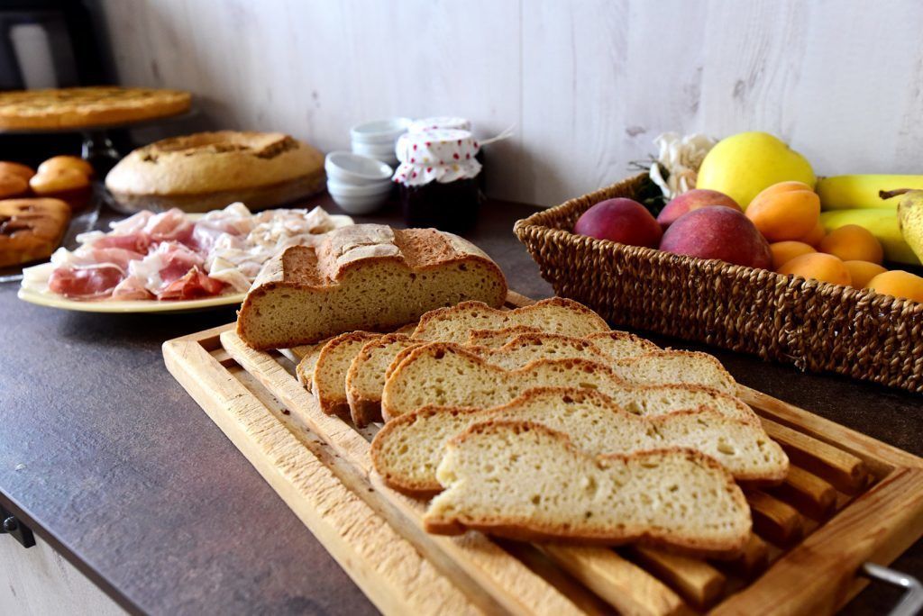 Tábua de madeira com pão fatiado, uma cesta de frutas e um prato de embutidos em uma bancada de cozinha.
