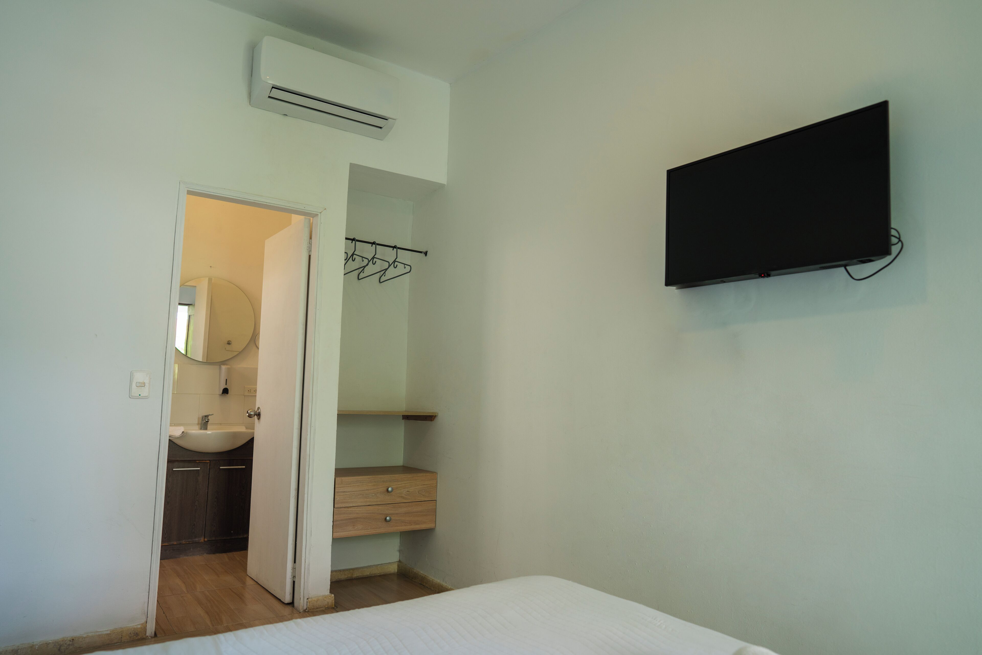 Quarto com cama, televisão na parede, guarda-roupa e banheiro visível através de uma porta aberta.