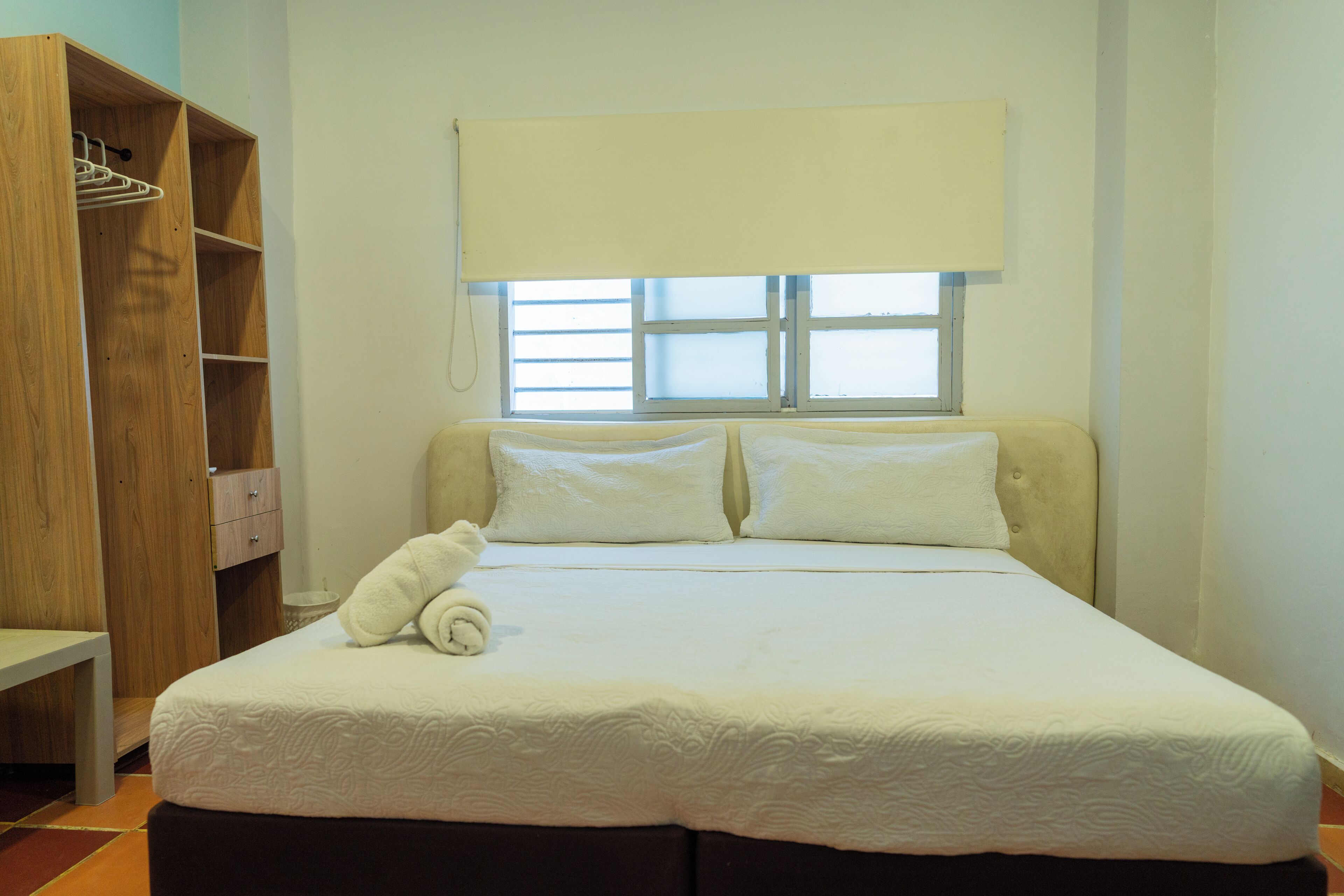 Cama bem arrumada com edredom branco, guarda-roupa de madeira e janela com persianas.