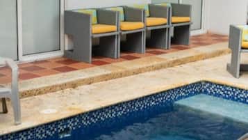 Uma área de piscina com borda em azulejo, um banco com almofadas e uma cadeira.
