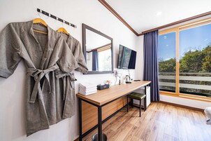 Quarto de hotel com escrivaninha de madeira, televisão, espelho e vista para o verde pela janela.