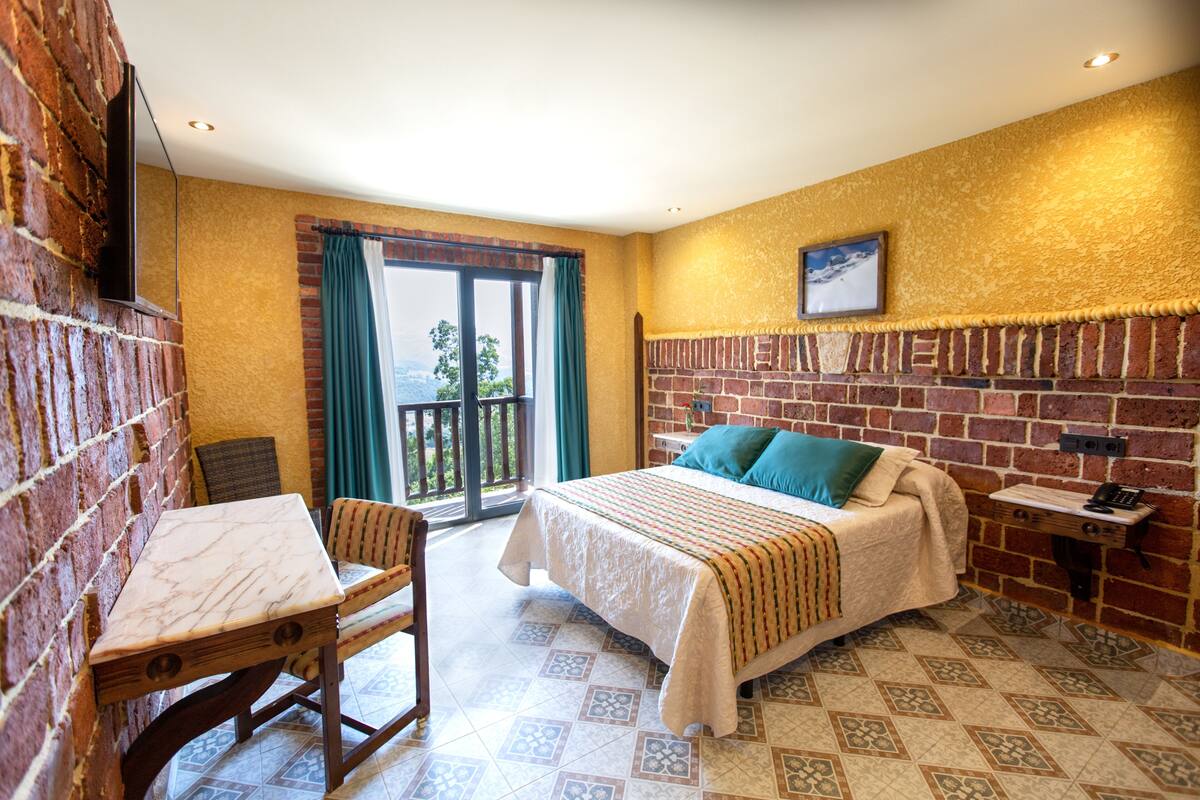 Quarto de hotel com cama, escrivaninha, cadeira, janela com cortinas e vista para o exterior.