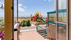 Vista de uma varanda com vista para um parquinho com escorregadores e estruturas de escalada, céu claro e uma praia ao fundo.