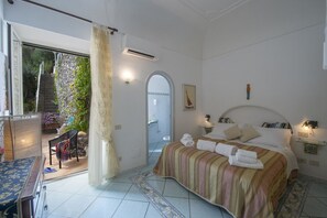 Quarto com uma cama grande, janela com cortinas, uma mesinha e uma cadeira, e vista para um jardim.