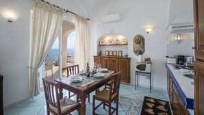 Uma sala de jantar com uma mesa posta para quatro pessoas, uma cozinha com micro-ondas e torradeira, e vista para o mar através de uma janela.