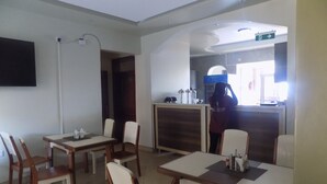 Uma sala de jantar de hotel com várias mesas e cadeiras, uma área de cozinha ao fundo, e uma pessoa em pé perto da cozinha.