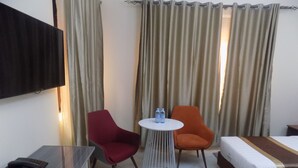 Quarto de hotel com uma cama, duas cadeiras, uma mesa pequena com garrafas de água, uma escrivaninha com telefone e cortinas.