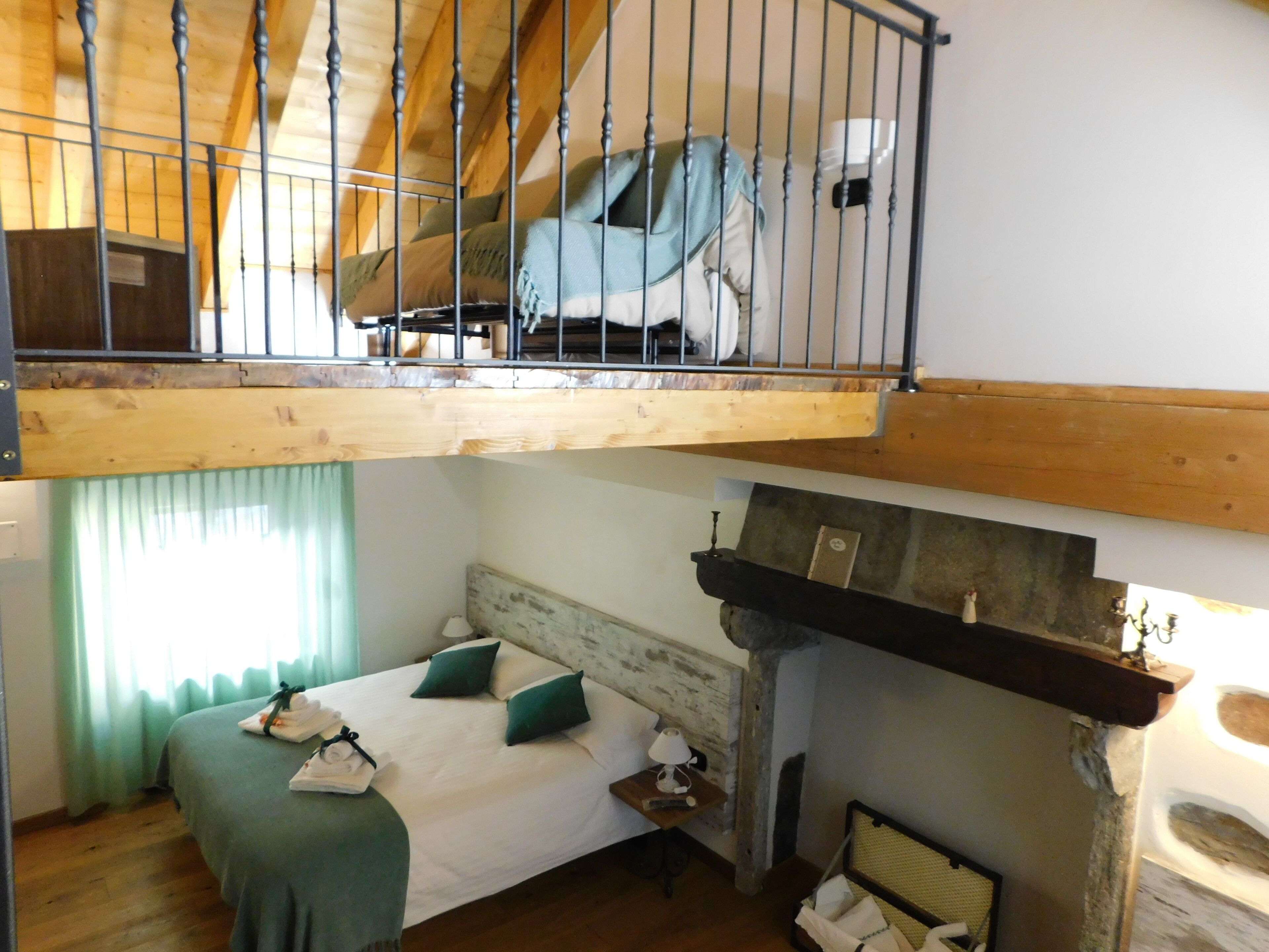 Quarto com beliche, uma cama de solteiro, vigas de madeira, janela com cortinas, mesa de cabeceira com luminária e uma pequena prateleira.