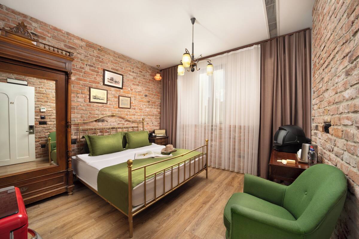 Quarto de hotel com parede de tijolos, cama de madeira, poltrona verde, televisão e minibar.