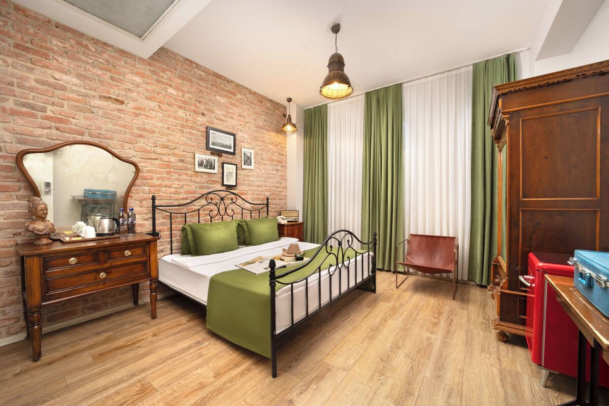 Quarto com parede de tijolos, cama com roupa de cama verde, cômoda, cadeira e eletrodoméstico vermelho antigo.
