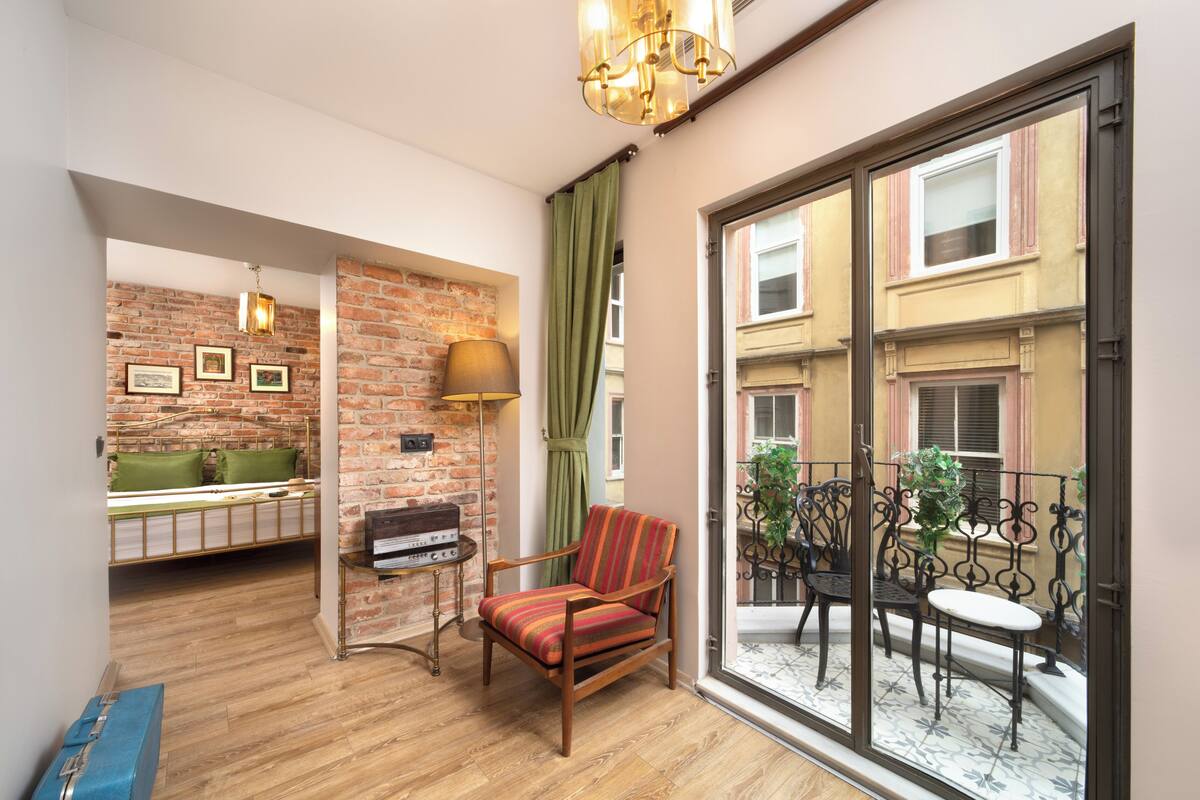 Um quarto acolhedor com parede de tijolos, piso de madeira, cortina verde, cadeira de madeira, mesa pequena com abajur e varanda com mobiliário de exterior.