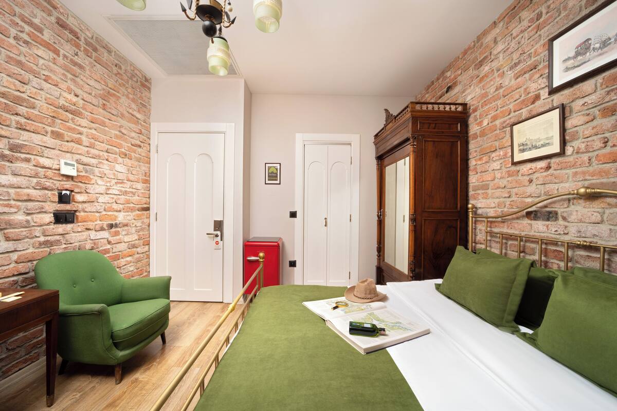 Um quarto com parede de tijolos, uma poltrona verde, uma cama com roupa de cama verde, um guarda-roupa de madeira e um armário vermelho.