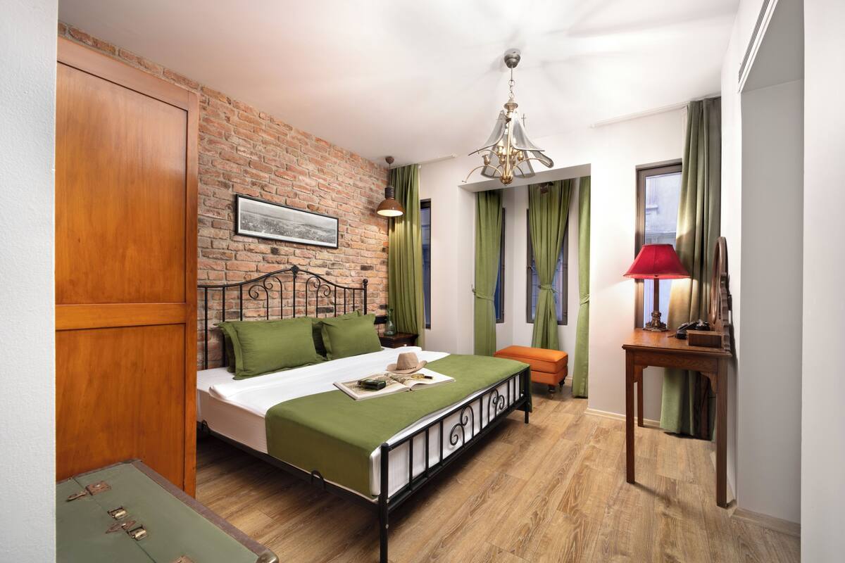 Quarto com parede de tijolos, cama com roupa de cama verde, guarda-roupa de madeira, escrivaninha com abajur e janela com cortinas.