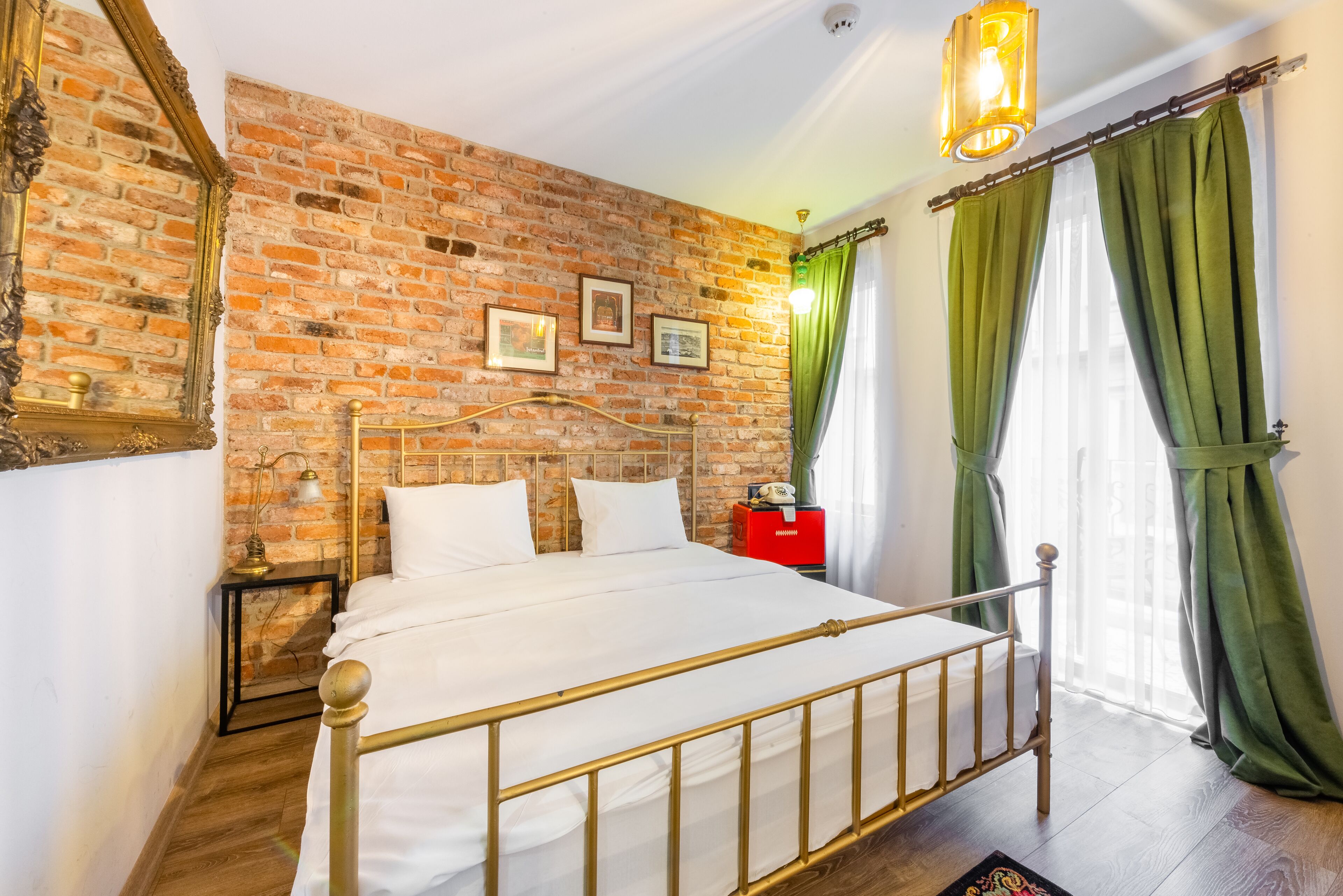 Um quarto com parede de tijolos, uma cama com estrutura metálica, uma mesa de cabeceira, um abajur e uma caixa vermelha.