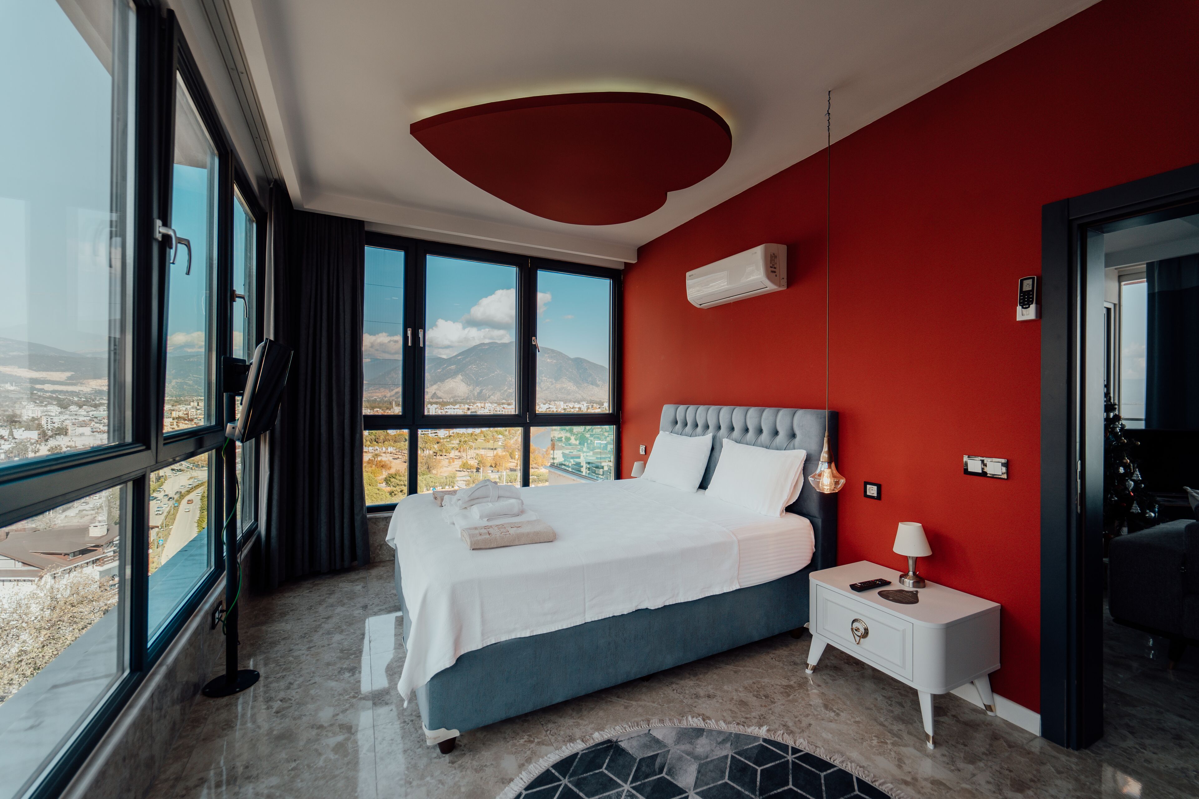Quarto moderno com uma cama grande, uma mesa de cabeceira e vista para a paisagem urbana através de grandes janelas.