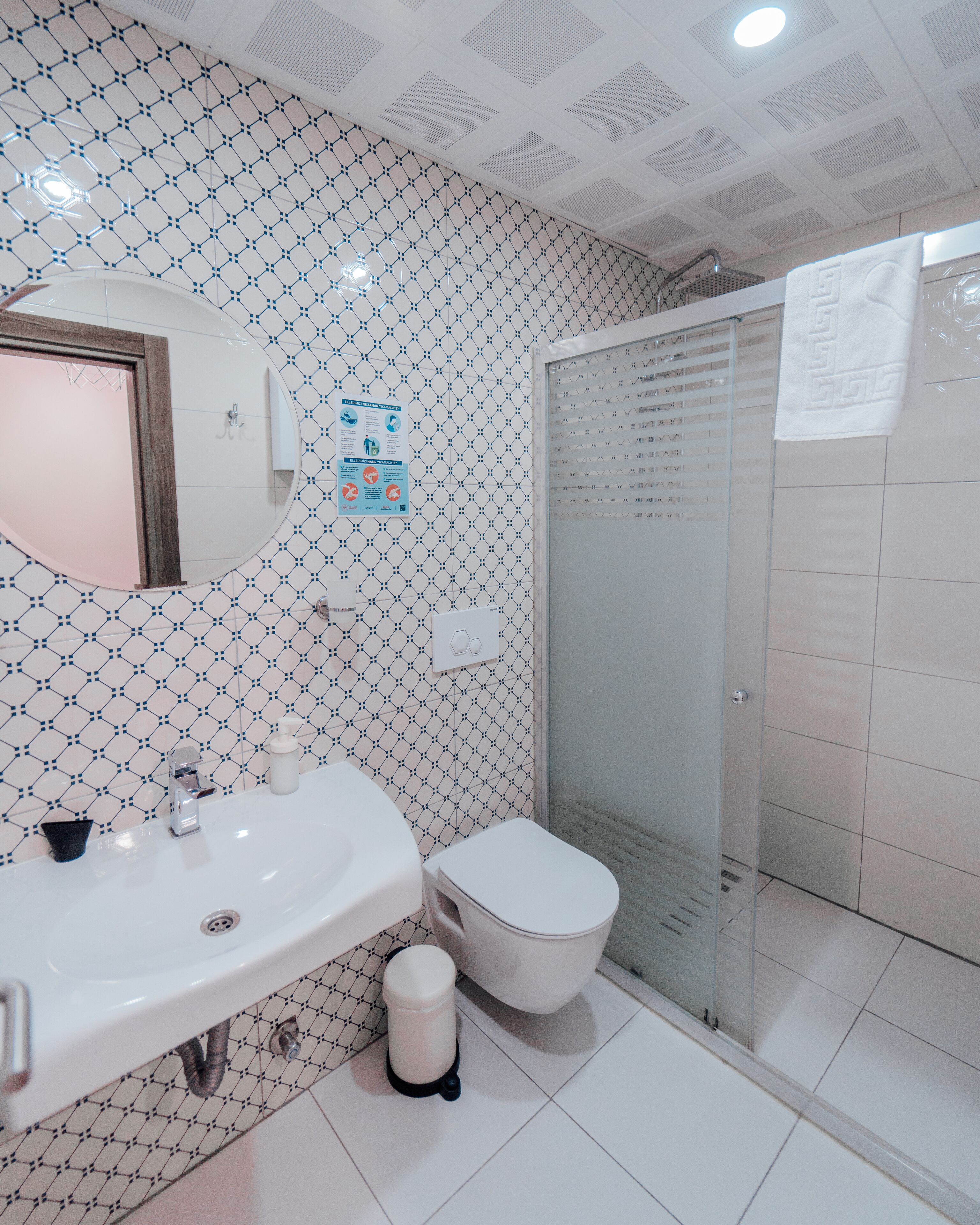 Casa de banho com lavatório branco, sanita e chuveiro. Há um espelho acima do lavatório e uma toalha pendurada na porta do chuveiro.