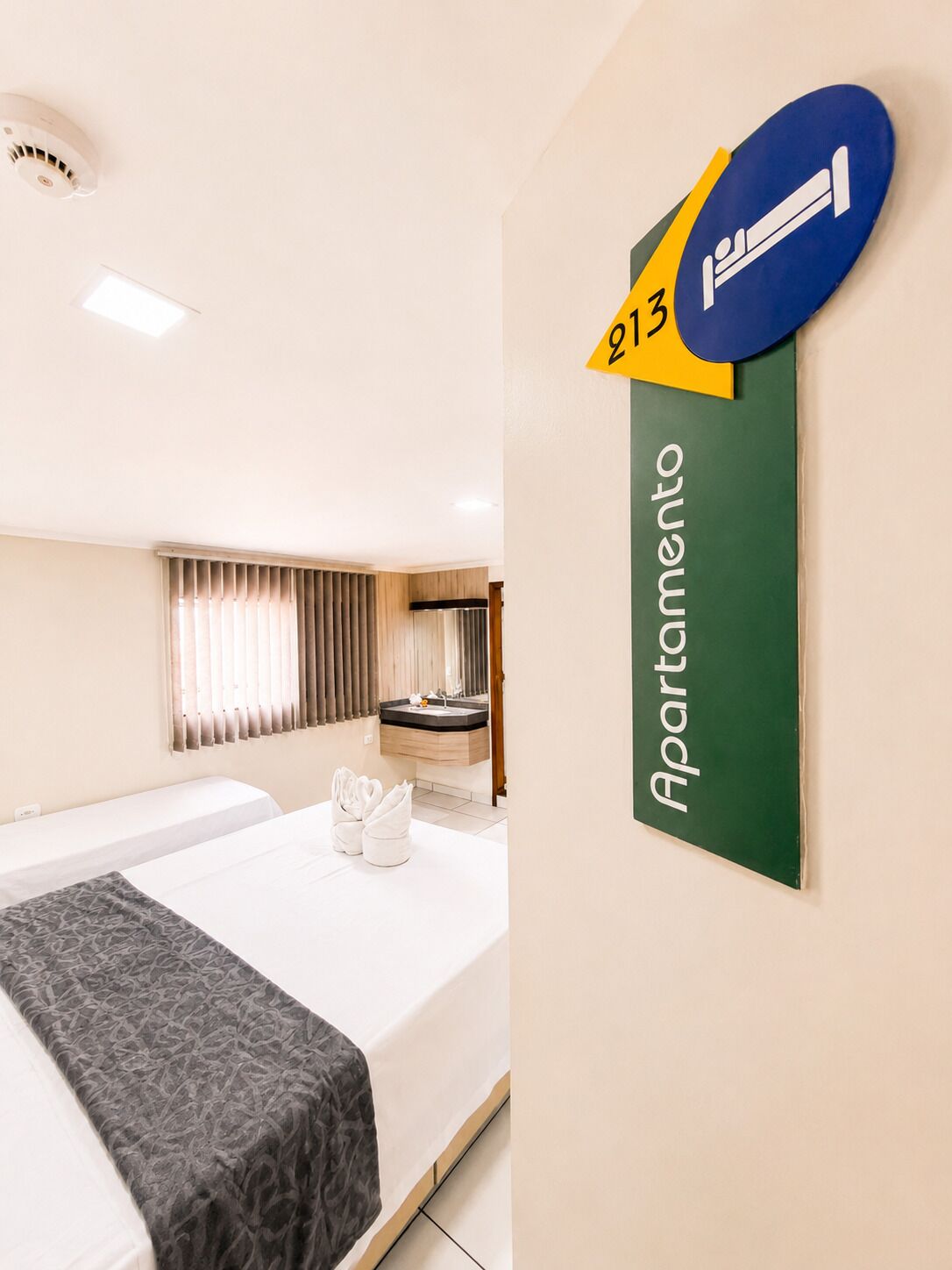 Quarto de hotel com cama, mesa de cabeceira, espelho, janela com persianas e placa na parede indicando o número e tipo do quarto.