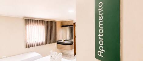 Quarto de hotel com cama, mesa de cabeceira, espelho, janela com persianas e placa na parede indicando o número e tipo do quarto.