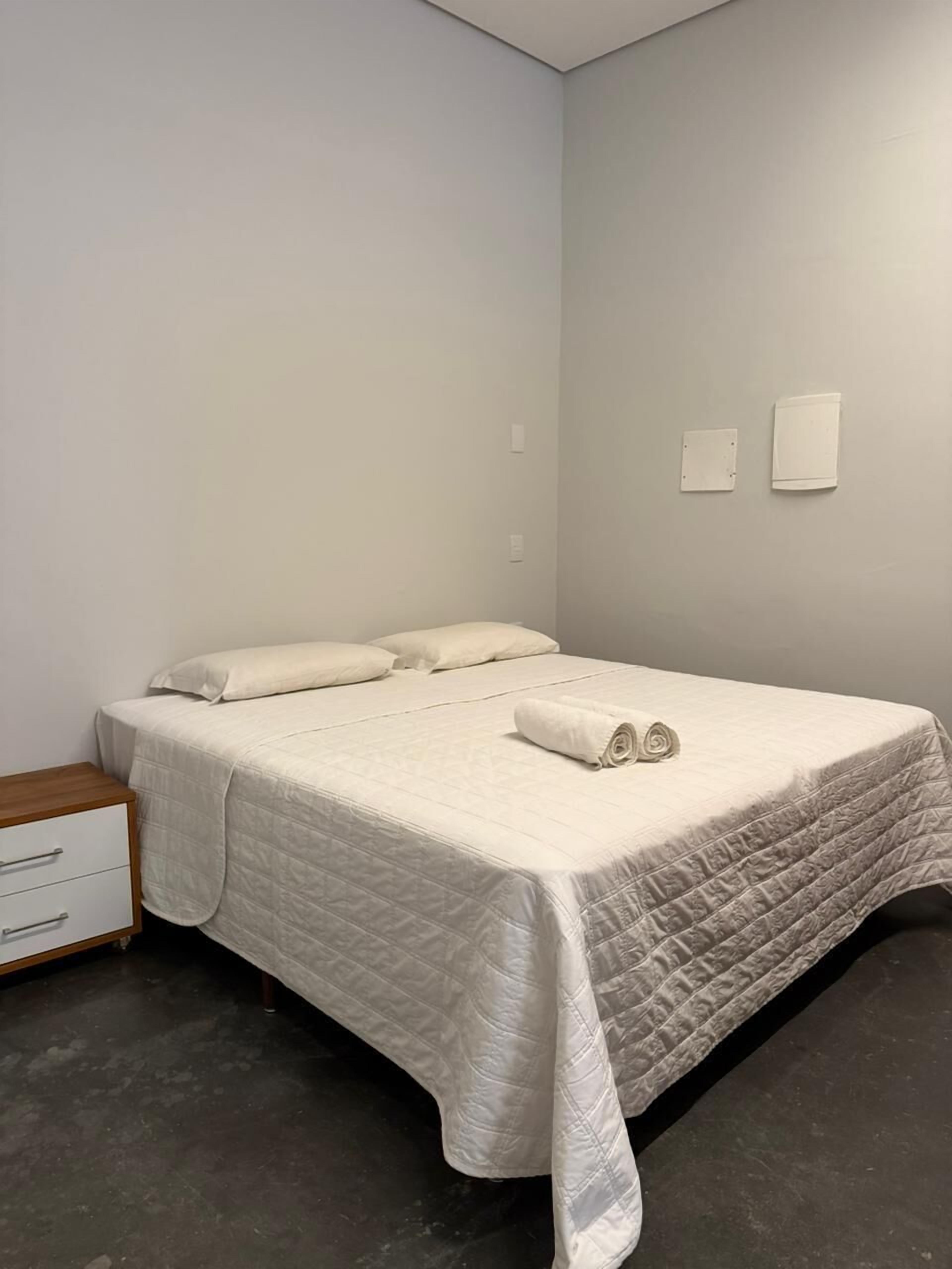 Uma cama de solteiro com colcha acolchoada, uma mesa de cabeceira de madeira com gavetas e dois interruptores de luz fixados na parede.