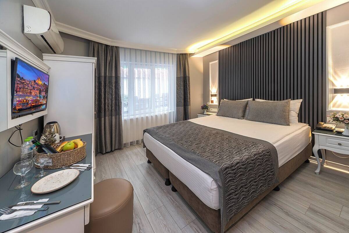 Quarto de hotel moderno com uma cama grande, uma escrivaninha com computador e uma televisão exibindo uma vista urbana, uma janela com cortinas e uma mesa de cabeceira com abajur.