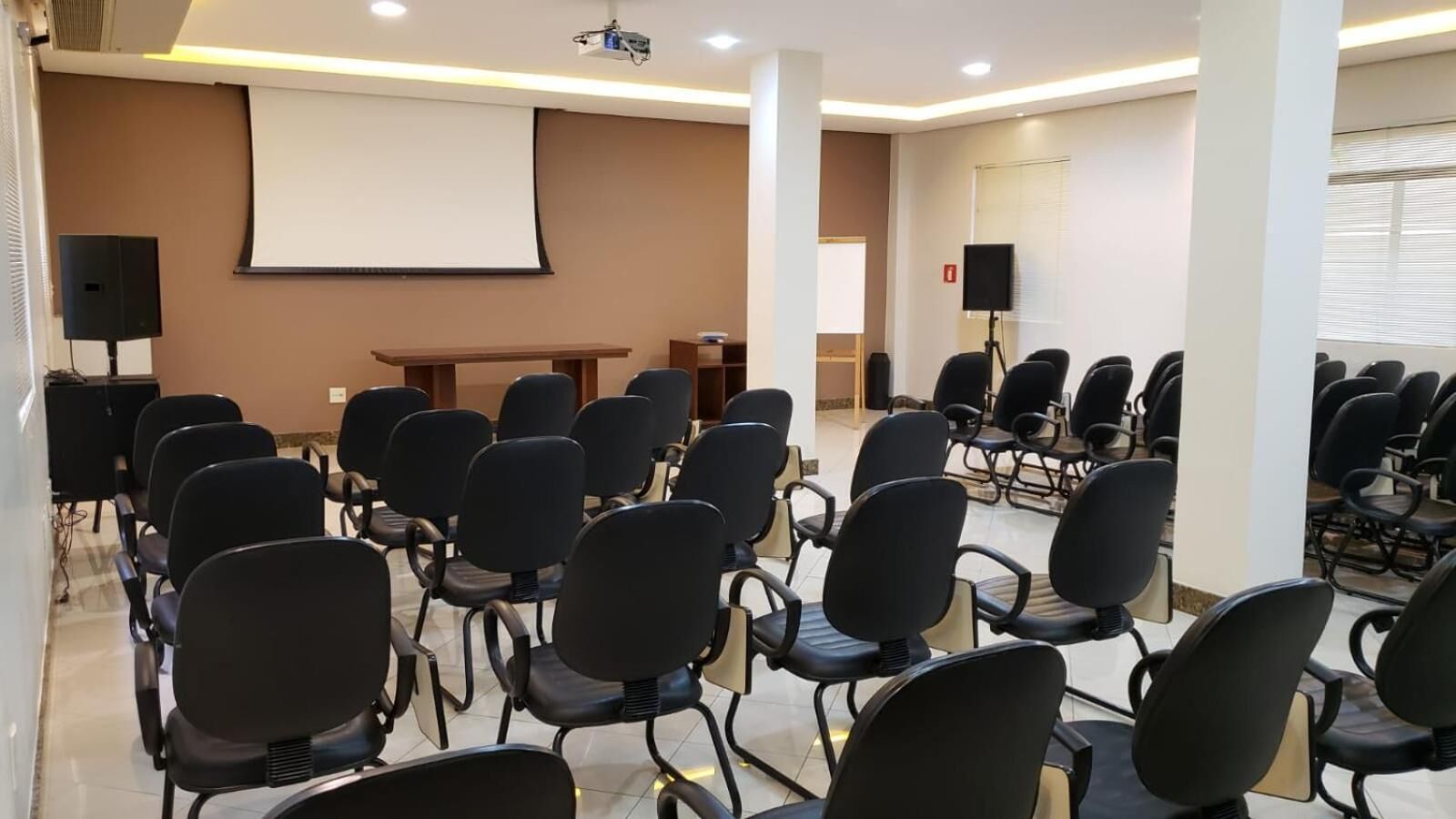 Uma sala de conferências com fileiras de cadeiras pretas, uma tela de projetor e um quadro branco.