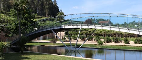 Uma ponte de vidro sobre um lago, com um gramado e árvores ao fundo.
