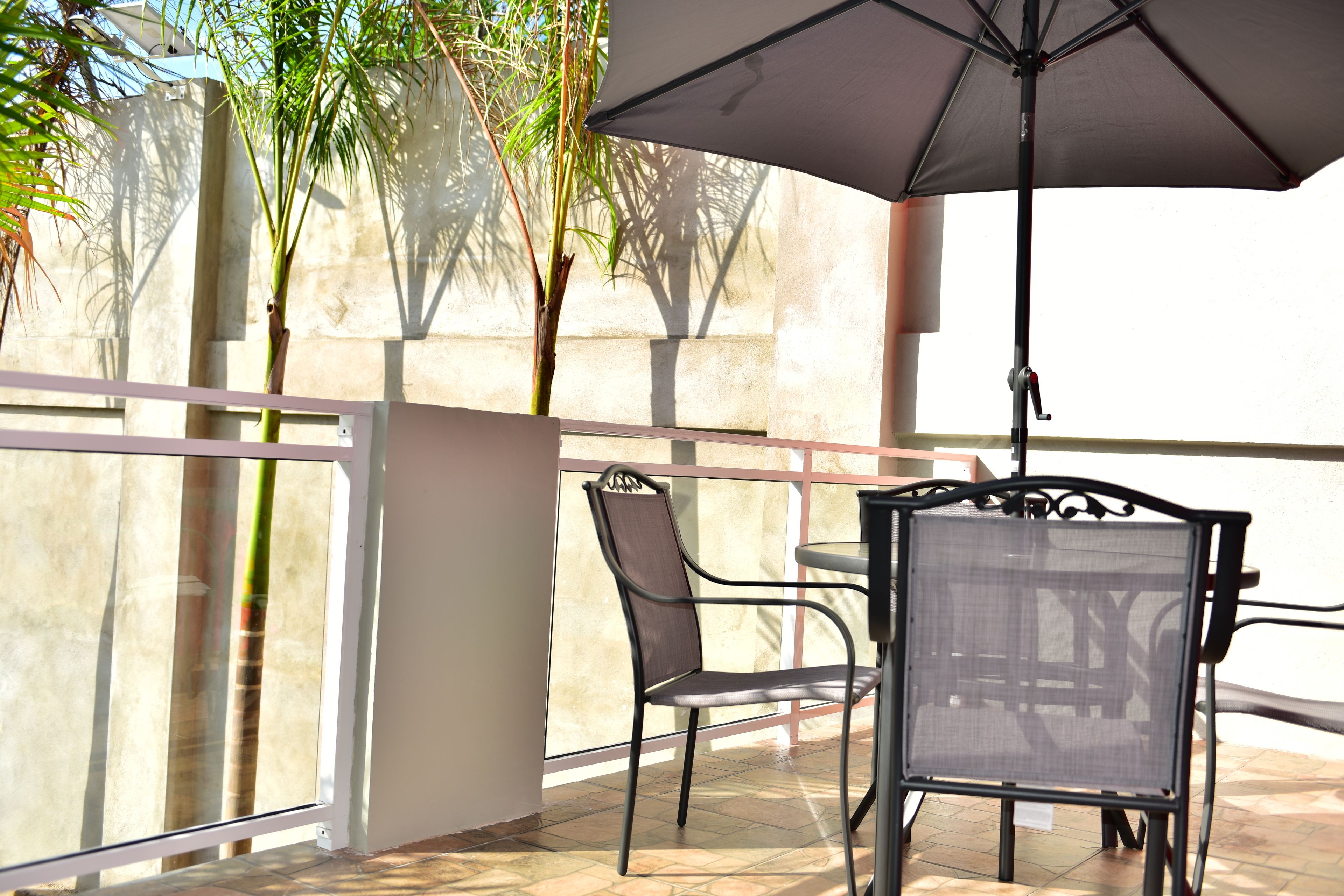 Varanda com mesa e cadeiras, um guarda-sol grande e vista para palmeiras e um edifício.