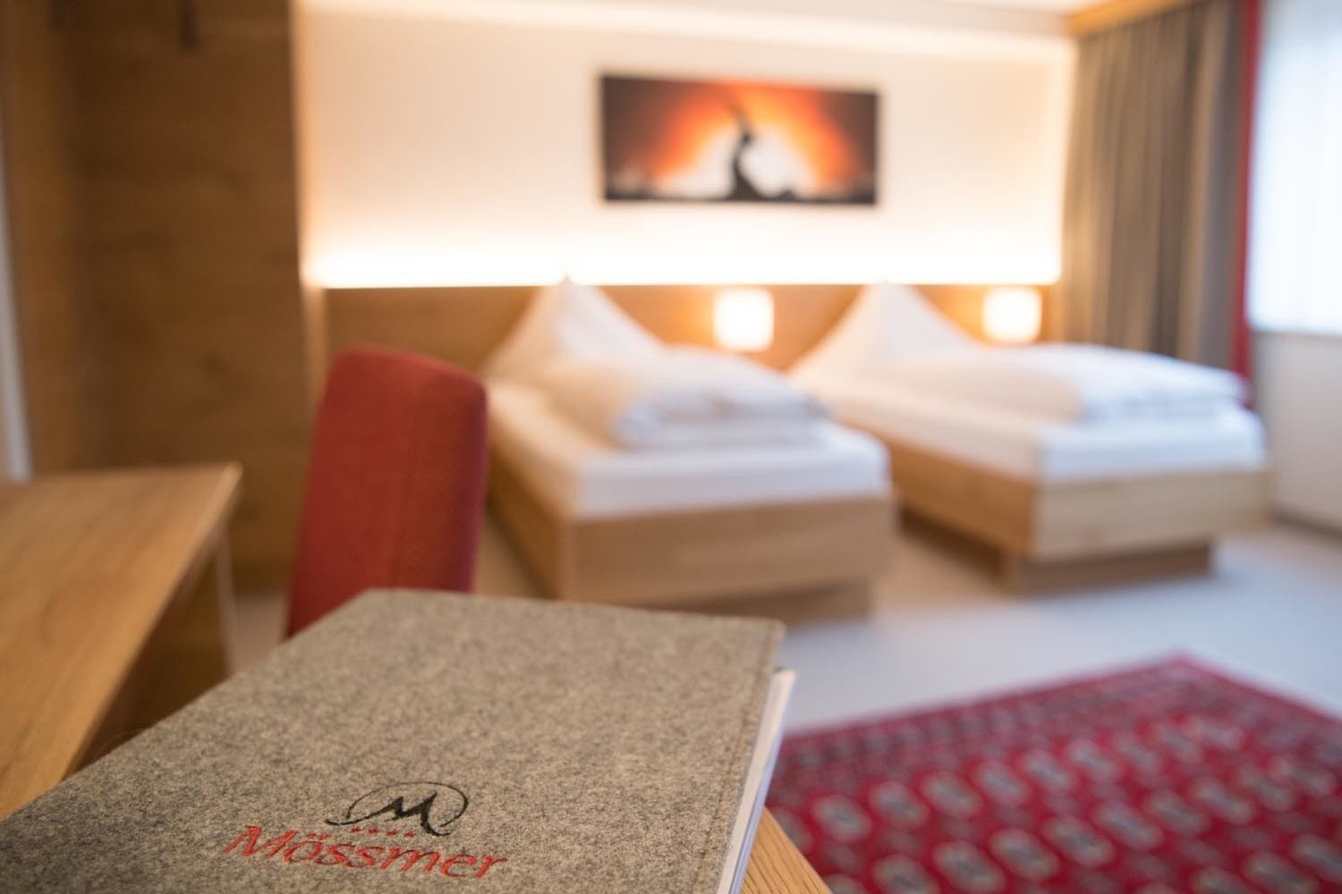 Quarto de hotel com uma cama, uma escrivaninha e uma cadeira. Há uma luminária na escrivaninha e um quadro na parede.