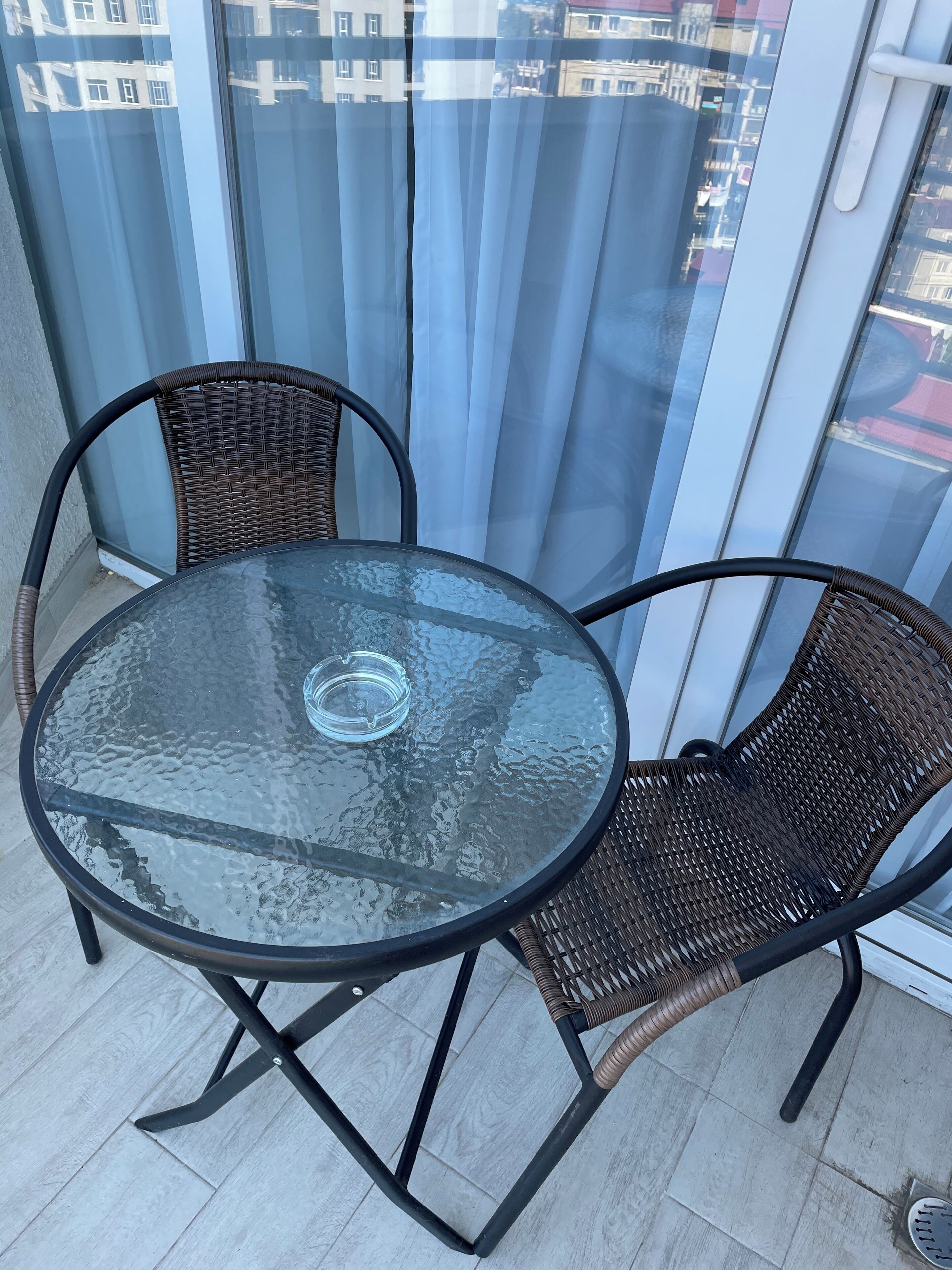 Uma mesa de vidro redonda com duas cadeiras de vime, colocada numa varanda com vista para edifícios através de cortinas leves.