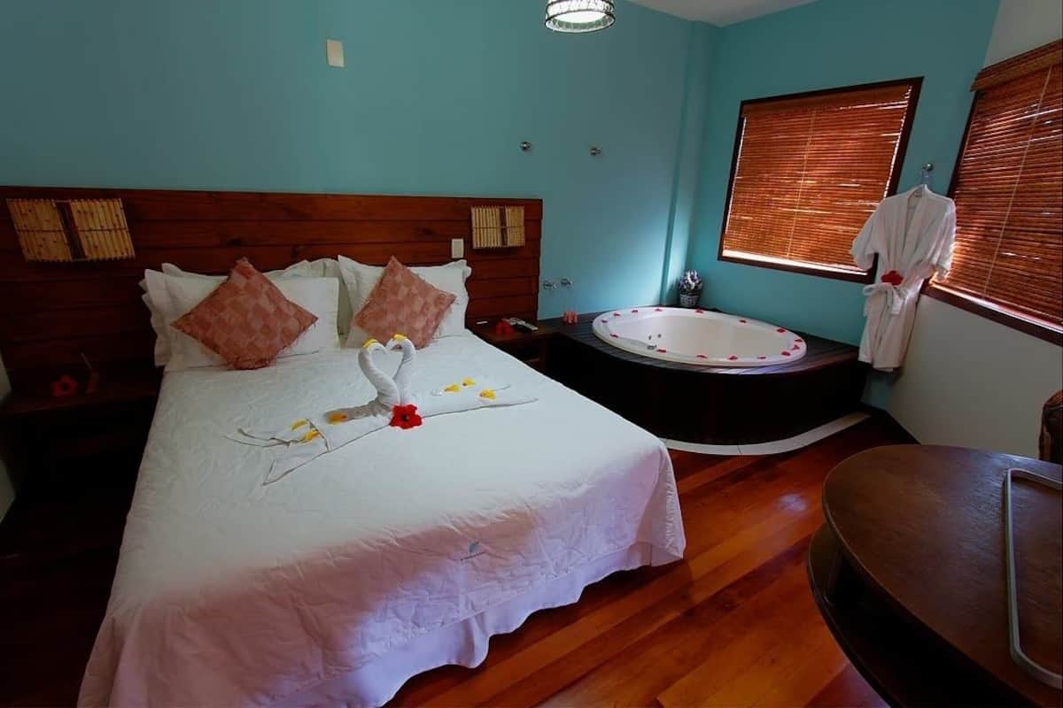 Um quarto com cabeceira de madeira, uma cama grande com roupa de cama branca, uma mesa de cabeceira, uma banheira embutida e uma janela com persianas.