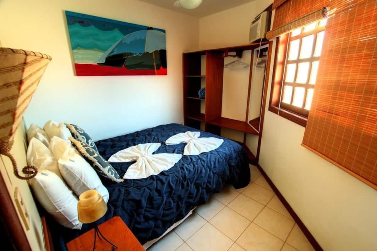 Quarto com cama, almofadas, um quadro, guarda-roupa e janela com persianas.