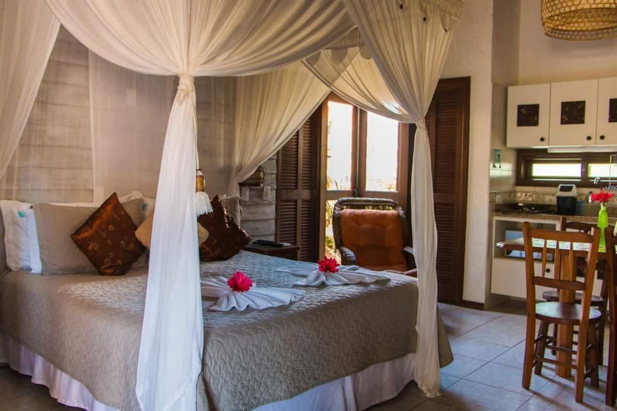 Quarto com cama de dossel, vista para cozinha e área de jantar com cadeiras.
