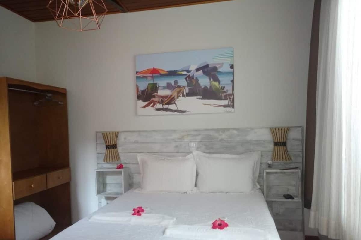 Quarto de hotel com uma cama, um quadro de uma cena de praia, um guarda-roupa de madeira e uma mesa de cabeceira com uma luminária.