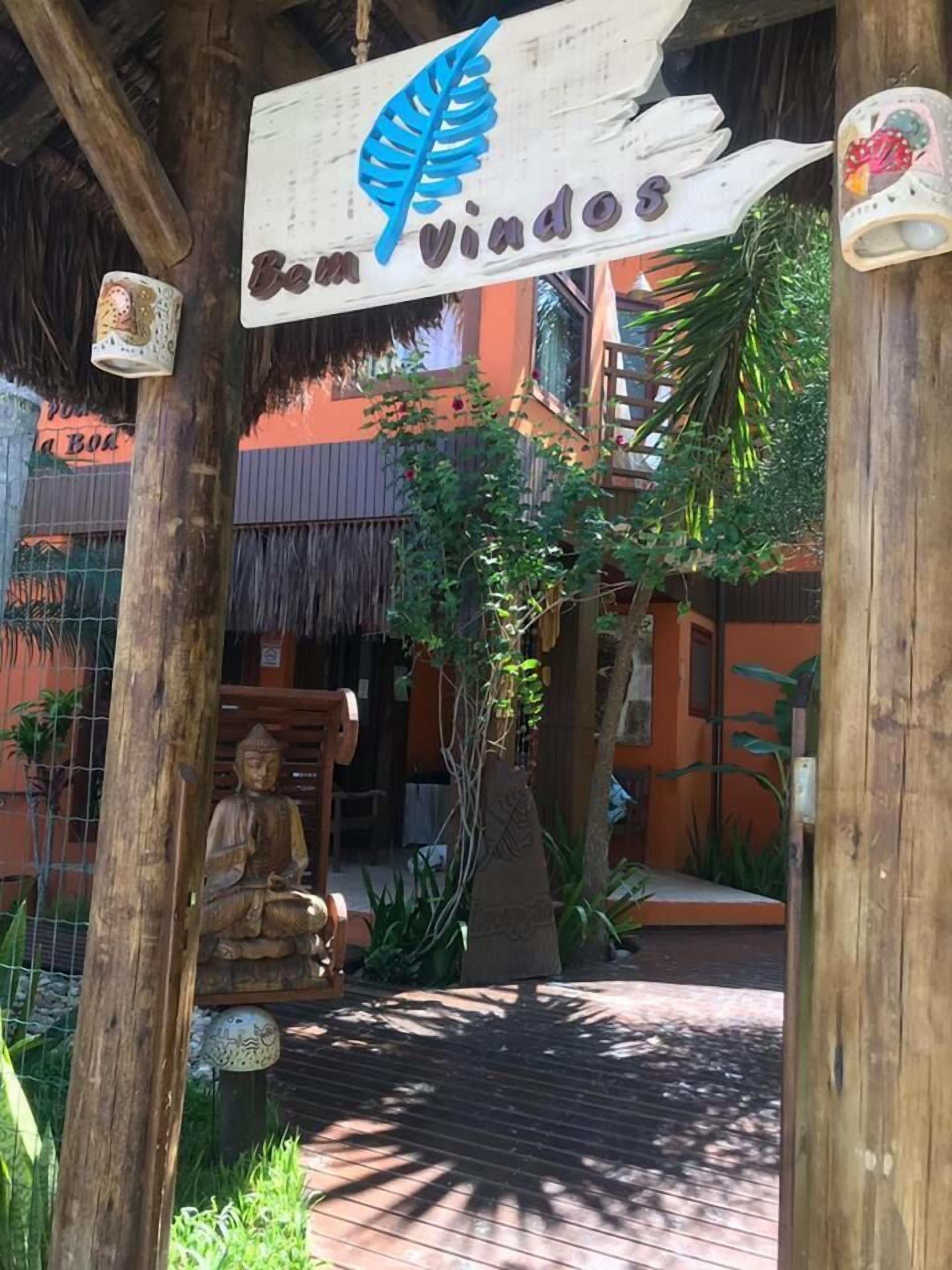 Placa de madeira com logotipo de folha e o texto 'Bom Vindo' sobre a entrada com telhado de palha.