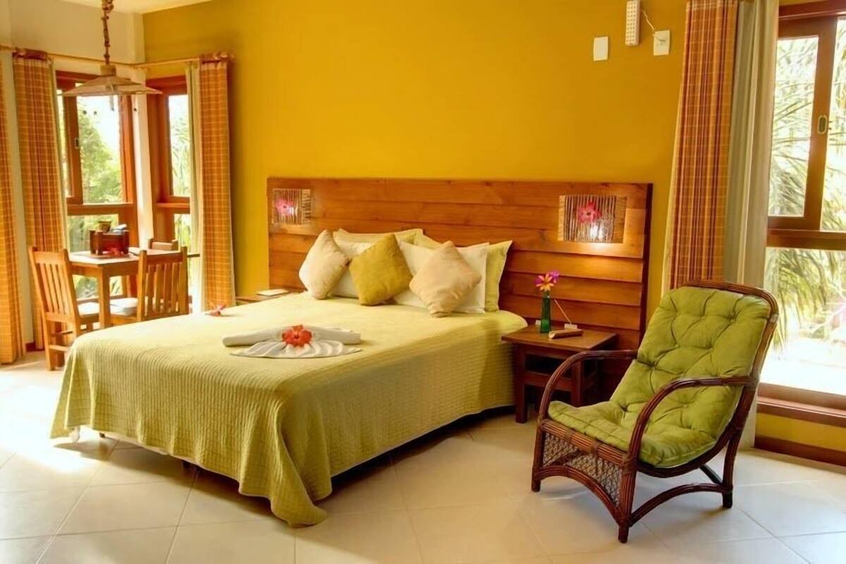 Quarto com cama, mesa de cabeceira, cadeira e janela com cortinas.