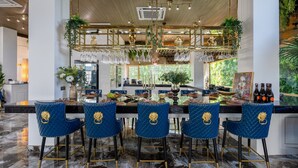 Uma área de bar com piso de mármore, banquetas azuis com detalhes dourados e uma vitrine de vidro.