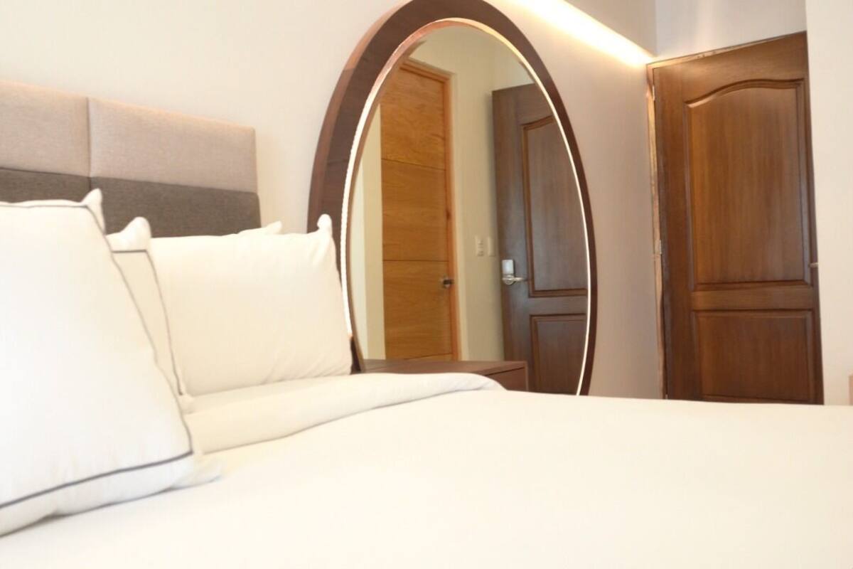 Cama bem arrumada com lençóis e almofadas brancas, porta de madeira e um espelho grande refletindo o quarto.
